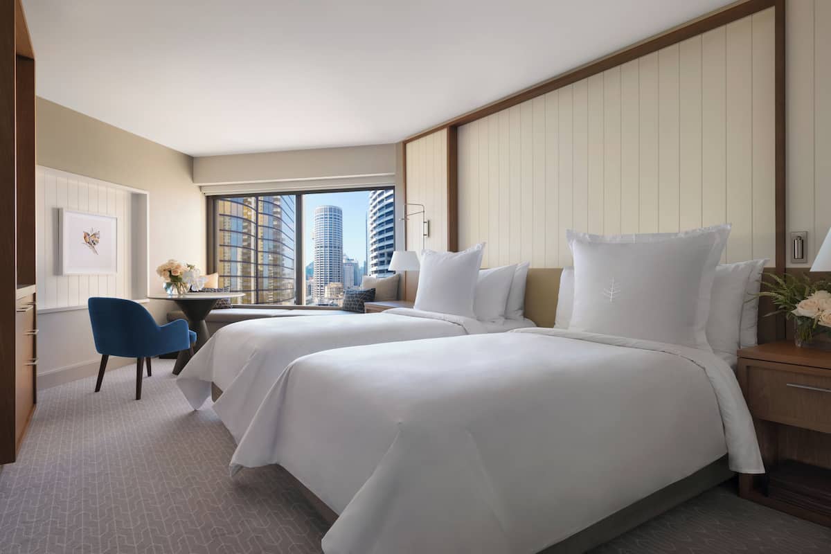 Quarto de hotel com duas camas, uma escrivaninha, uma cadeira e uma janela com vista para a cidade.
