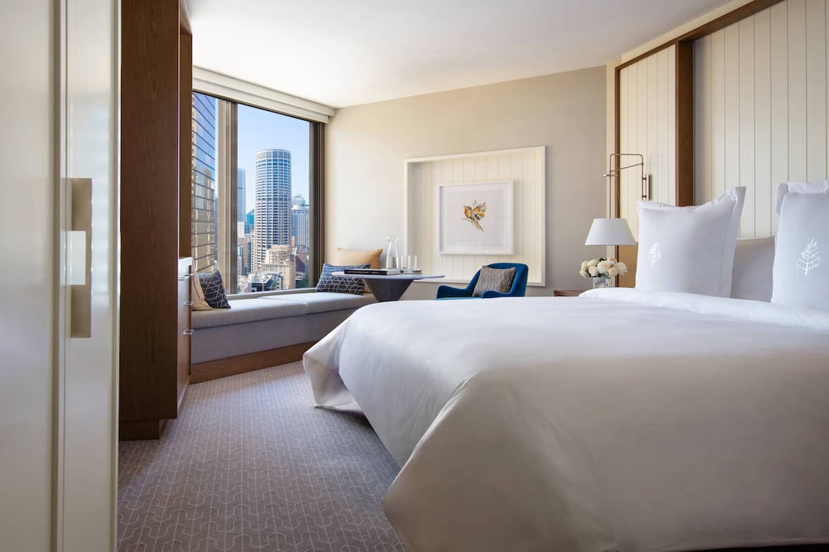 Quarto de hotel com uma cama grande, uma mesa de trabalho com cadeira, uma janela com vista para a cidade e um quadro na parede.