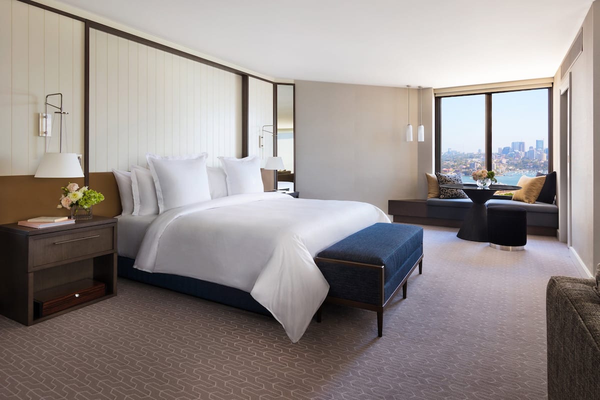 Quarto de hotel moderno com uma cama grande, área de estar com vista e paisagem urbana visível pela janela.