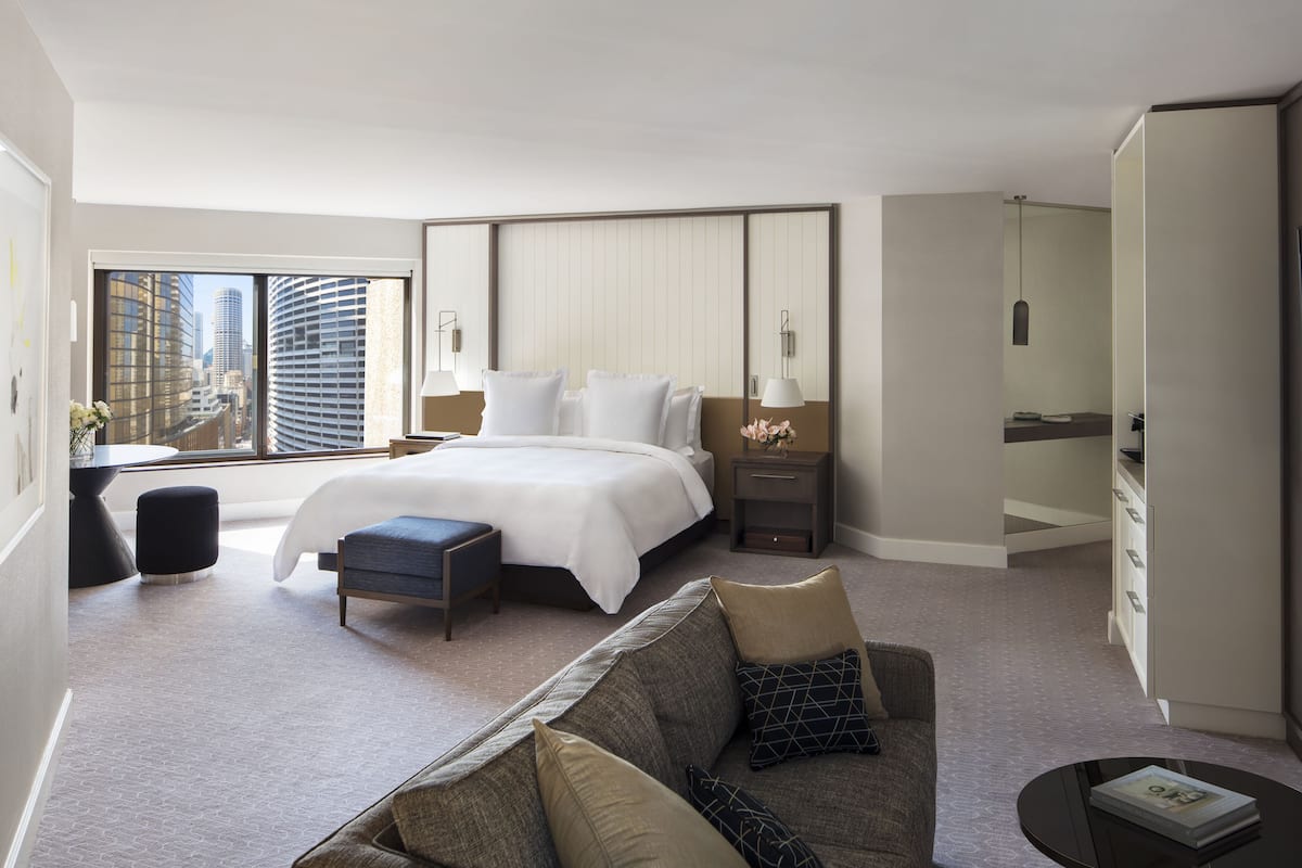 Quarto de hotel moderno com uma cama grande, um sofá, uma escrivaninha e vista da cidade através de janelas amplas.