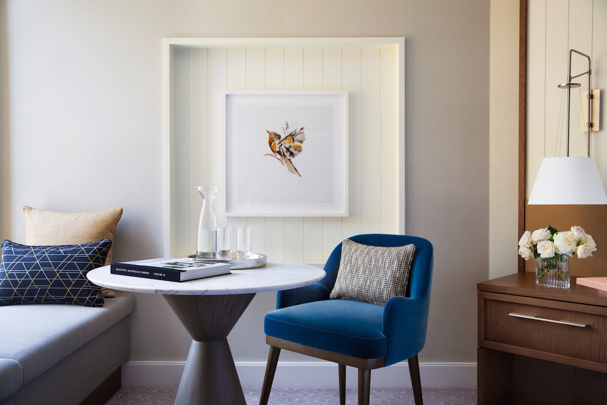 Quarto moderno com poltrona azul, mesa redonda pequena com um livro e copos, quadro pendurado na parede e mesa de cabeceira com abajur e vaso de flores.