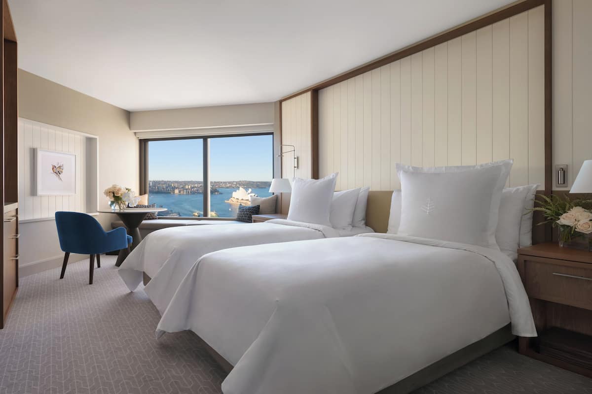 Quarto de hotel com duas camas, uma escrivaninha, uma cadeira e uma janela ampla com vista para a cidade.
