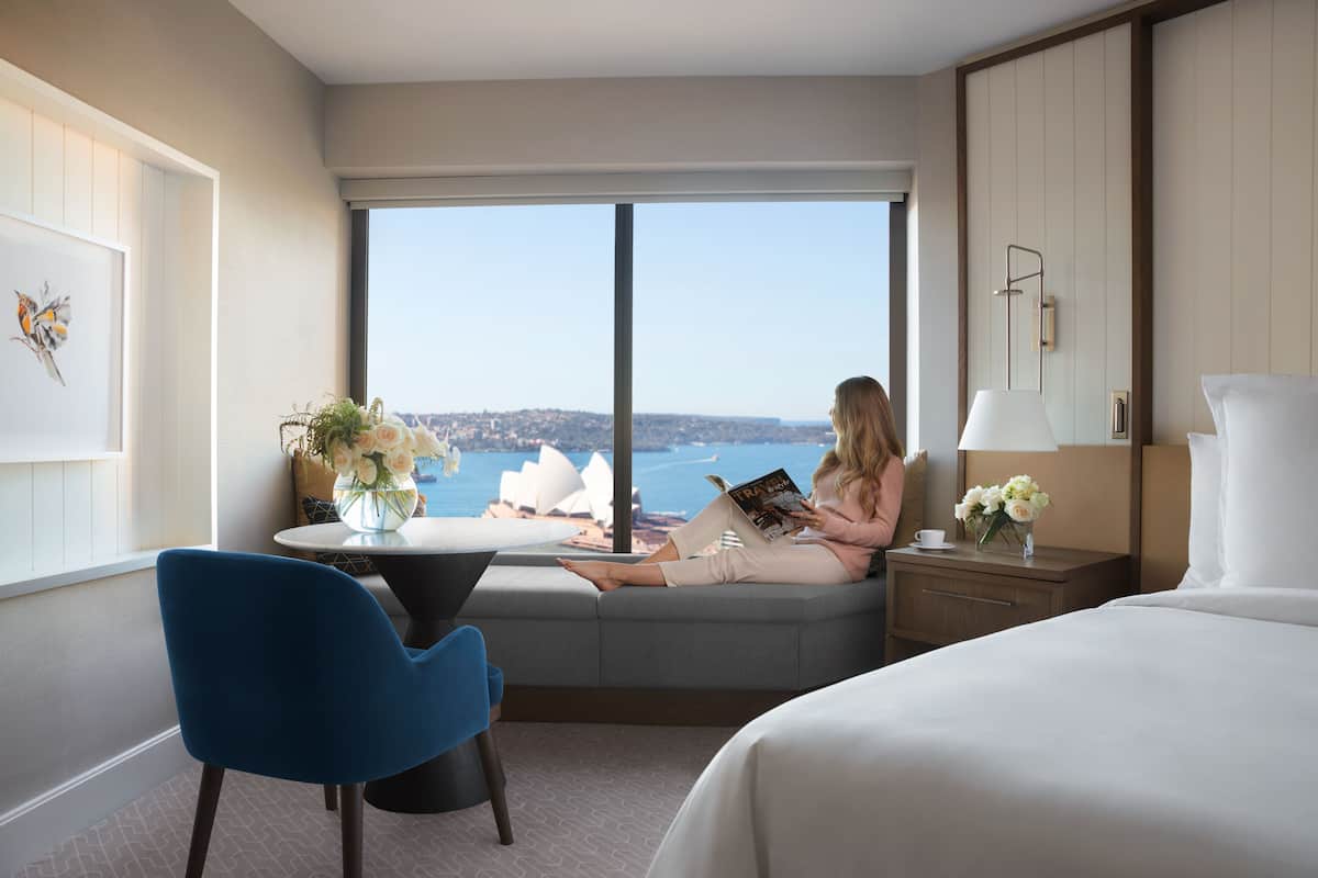 Quarto de hotel com uma janela grande, uma cama, uma mesa de cabeceira com abajur, uma cadeira e uma pequena mesa com um vaso de flores.