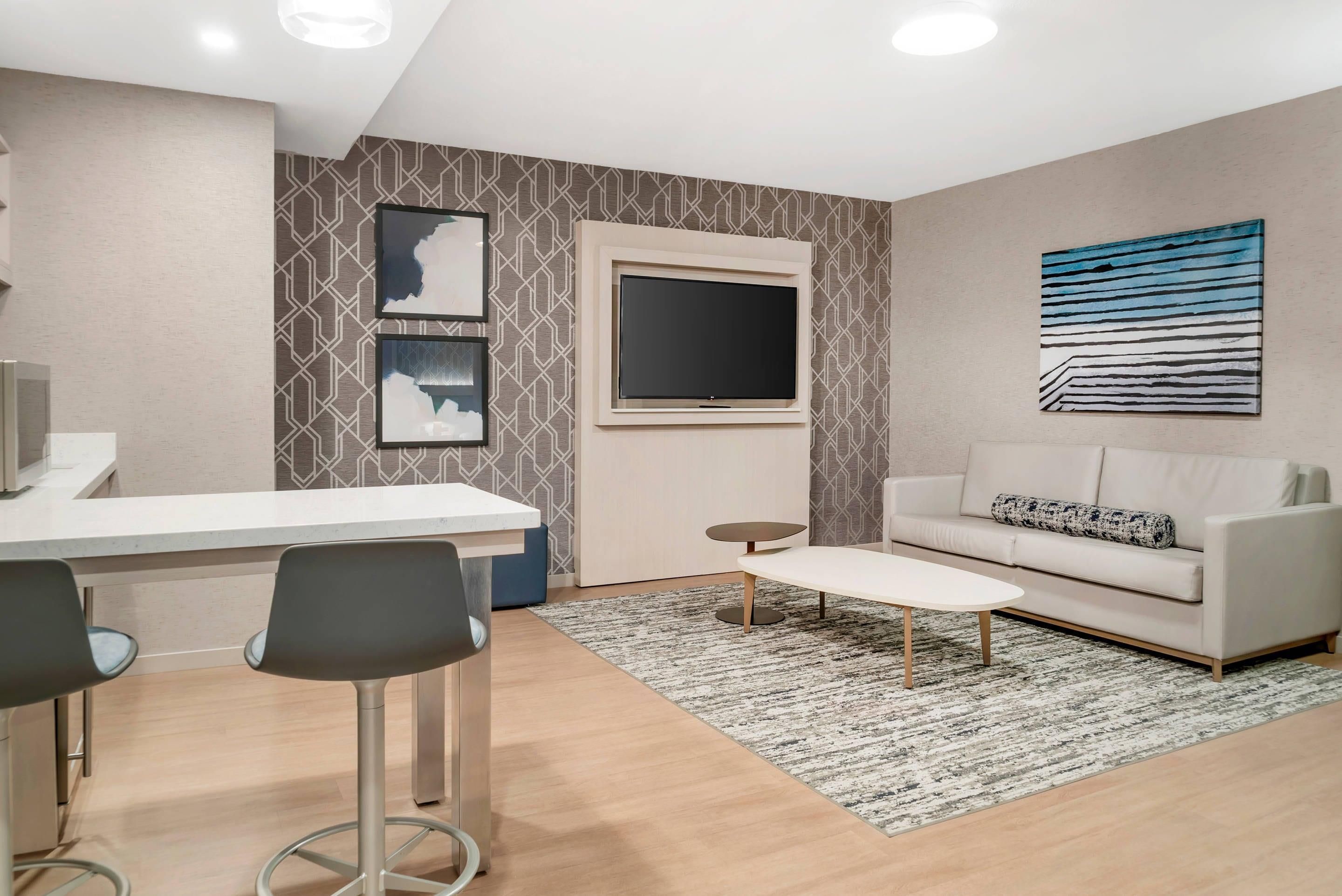 Uma sala de estar moderna com um sofá branco, uma mesa de centro e uma TV fixada na parede. Há dois bancos altos e uma pequena mesa redonda. O quarto possui papel de parede estampado e um tapete no chão.
