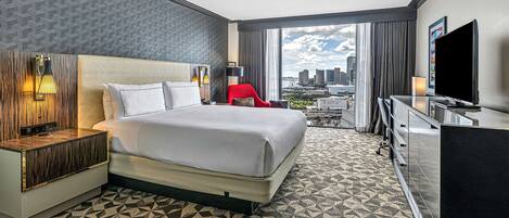 Quarto de hotel com uma cama grande, uma cadeira vermelha, uma escrivaninha com televisão e vista para a cidade pela janela.