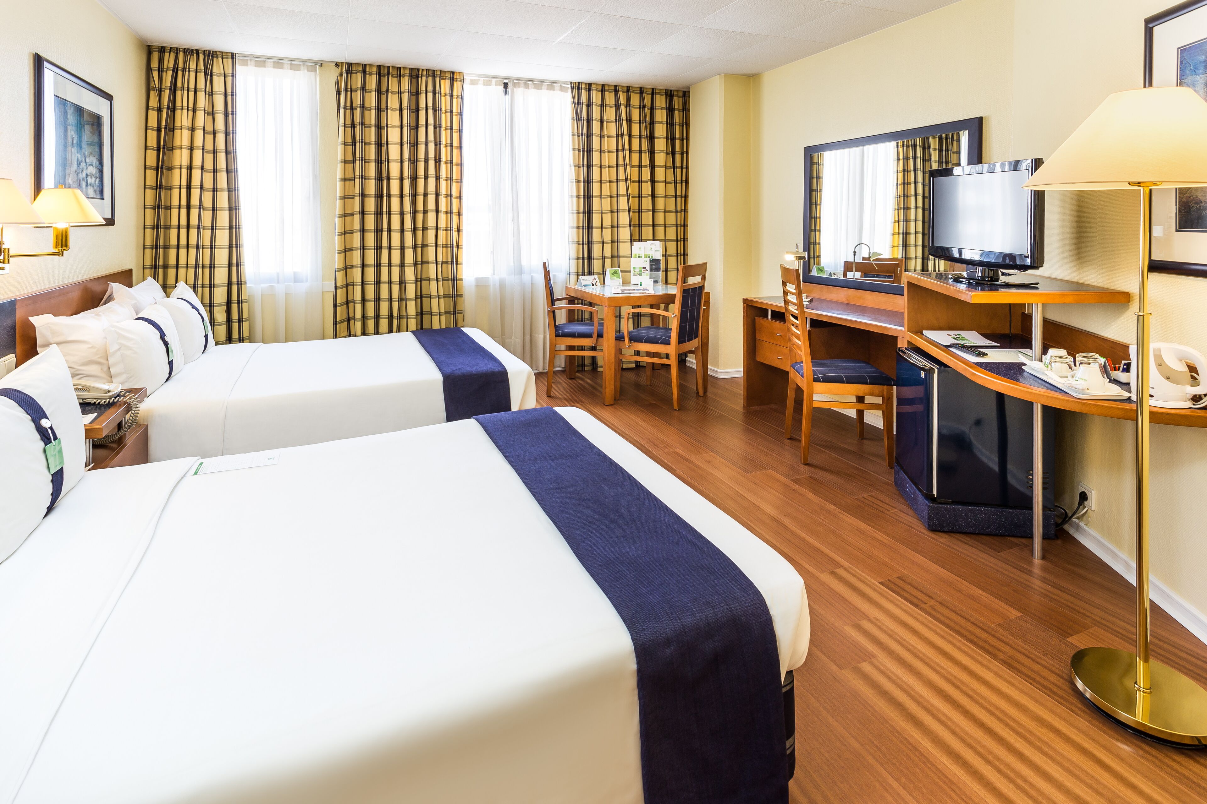 Quarto de hotel com duas camas, uma escrivaninha, uma cadeira, uma televisão e uma luminária de mesa.