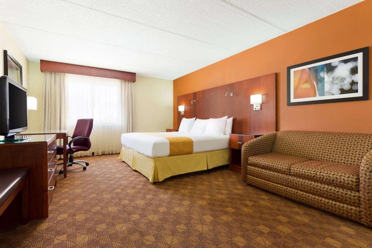 Quarto de hotel com cama, sofá, escrivaninha com televisão e janela com cortinas.