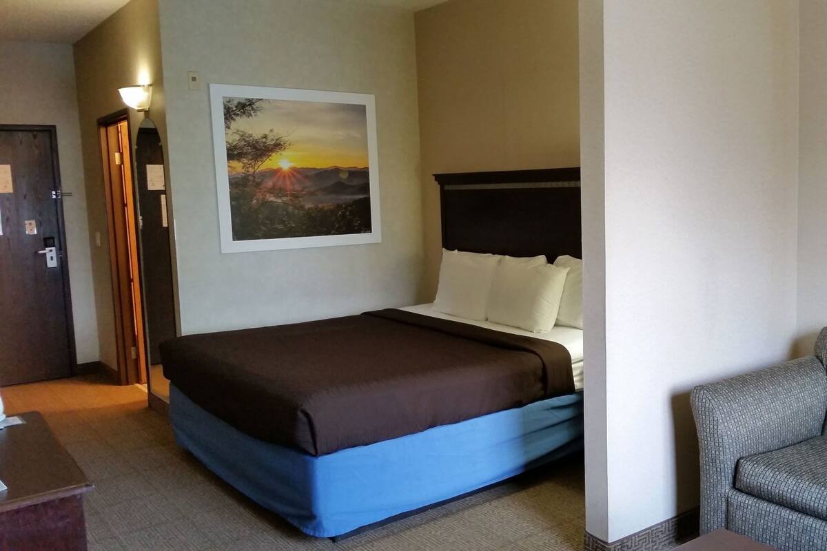 Quarto de hotel com uma cama, uma cadeira, uma escrivaninha e uma pintura de um pôr do sol.