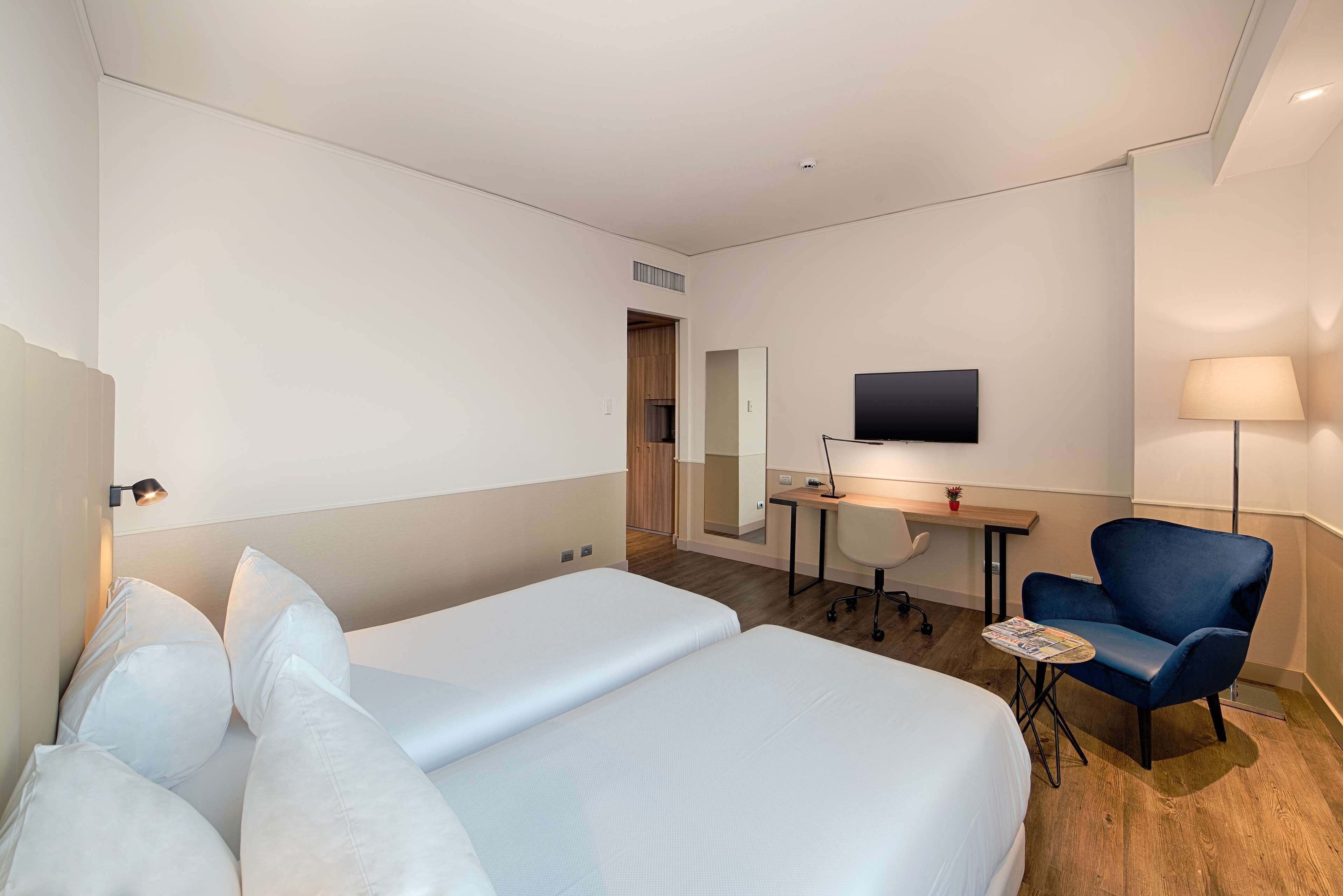 Quarto de hotel com duas camas, uma escrivaninha, uma cadeira, uma televisão e um abajur.