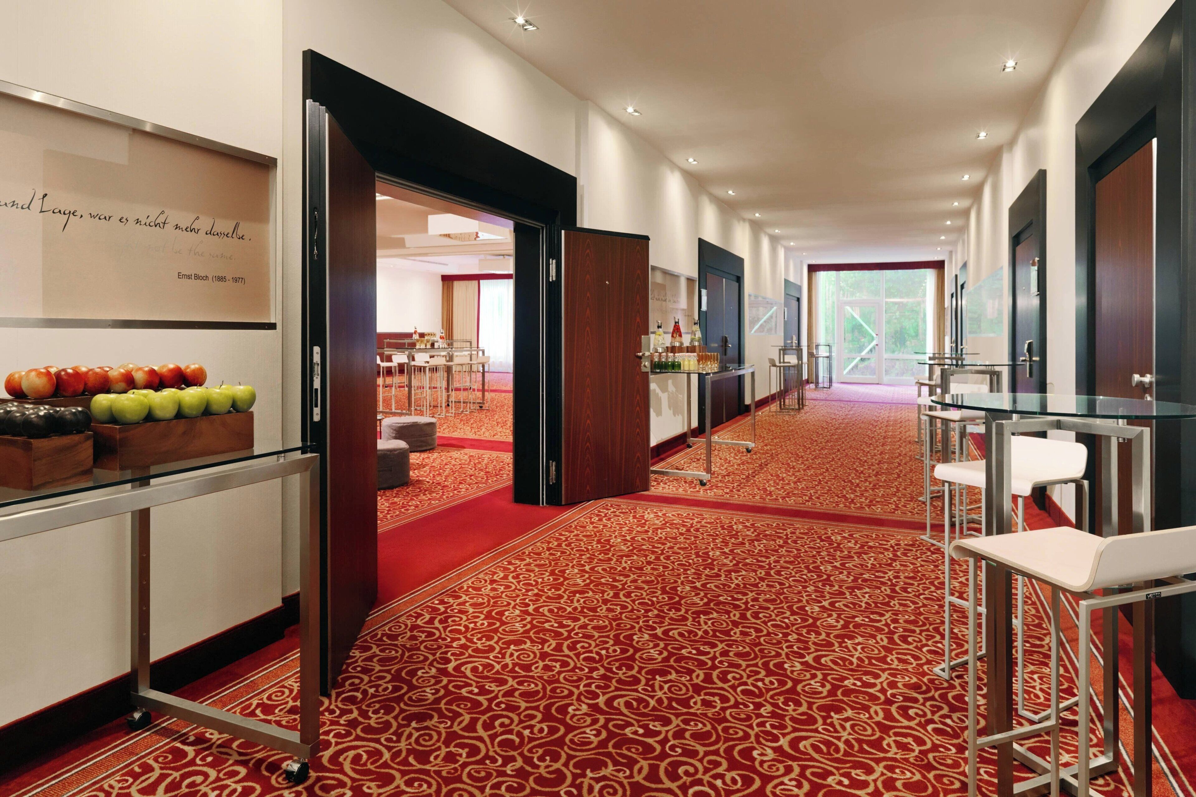 Corredor de hotel com carpete estampado em vermelho, área de bar com banquetas e uma parede com uma citação e maçãs.