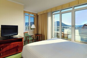 Quarto de hotel com cama, televisão, cadeira, abajur e grandes janelas com vista.