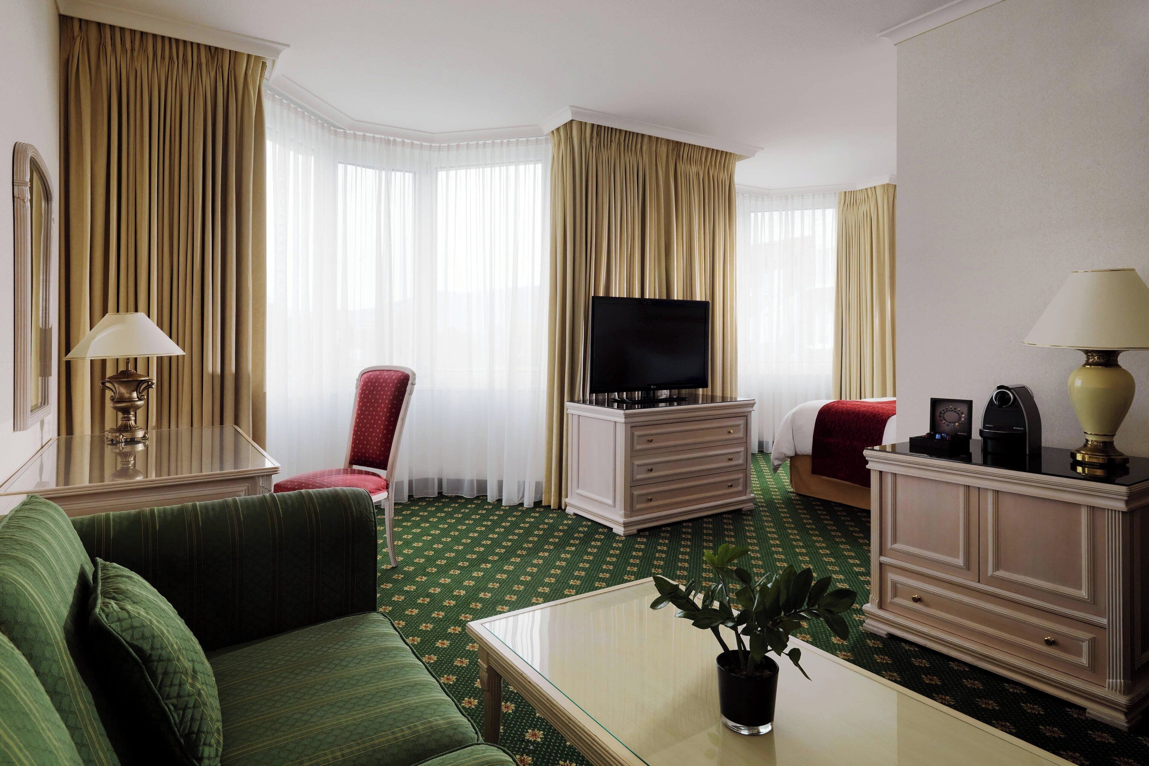 Quarto de hotel com carpete estampado verde, sofá verde, poltrona vermelha, televisão sobre um móvel e candeeiro num apoio lateral.