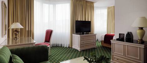 Quarto de hotel com carpete estampado verde, sofá verde, poltrona vermelha, televisão sobre um móvel e candeeiro num apoio lateral.