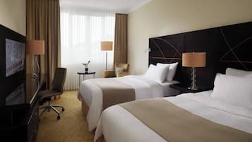 Quarto de hotel com duas camas, uma mesa de trabalho com televisão, uma cadeira, abajur e janela com cortinas.