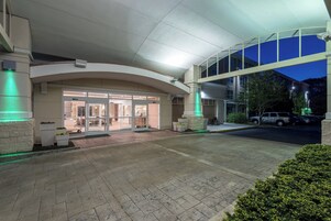 Entrada de hotel com galeria coberta, portas de vidro e área de estacionamento.