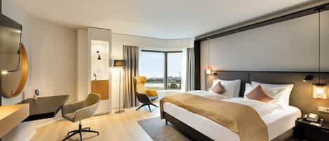 Quarto de hotel moderno com uma cama grande, uma escrivaninha, uma cadeira e vista para a cidade através de amplas janelas.