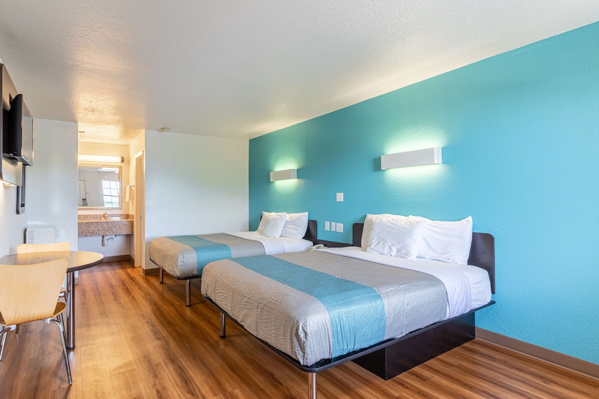 Quarto de hotel com duas camas, uma mesa pequena e uma cadeira. Há uma kitchenette ao fundo.