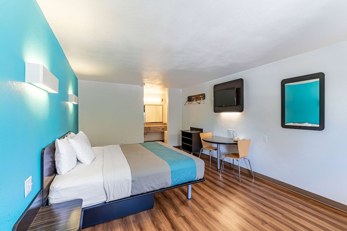 Quarto de hotel compacto com cama, escrivaninha, cadeira e televisão na parede.