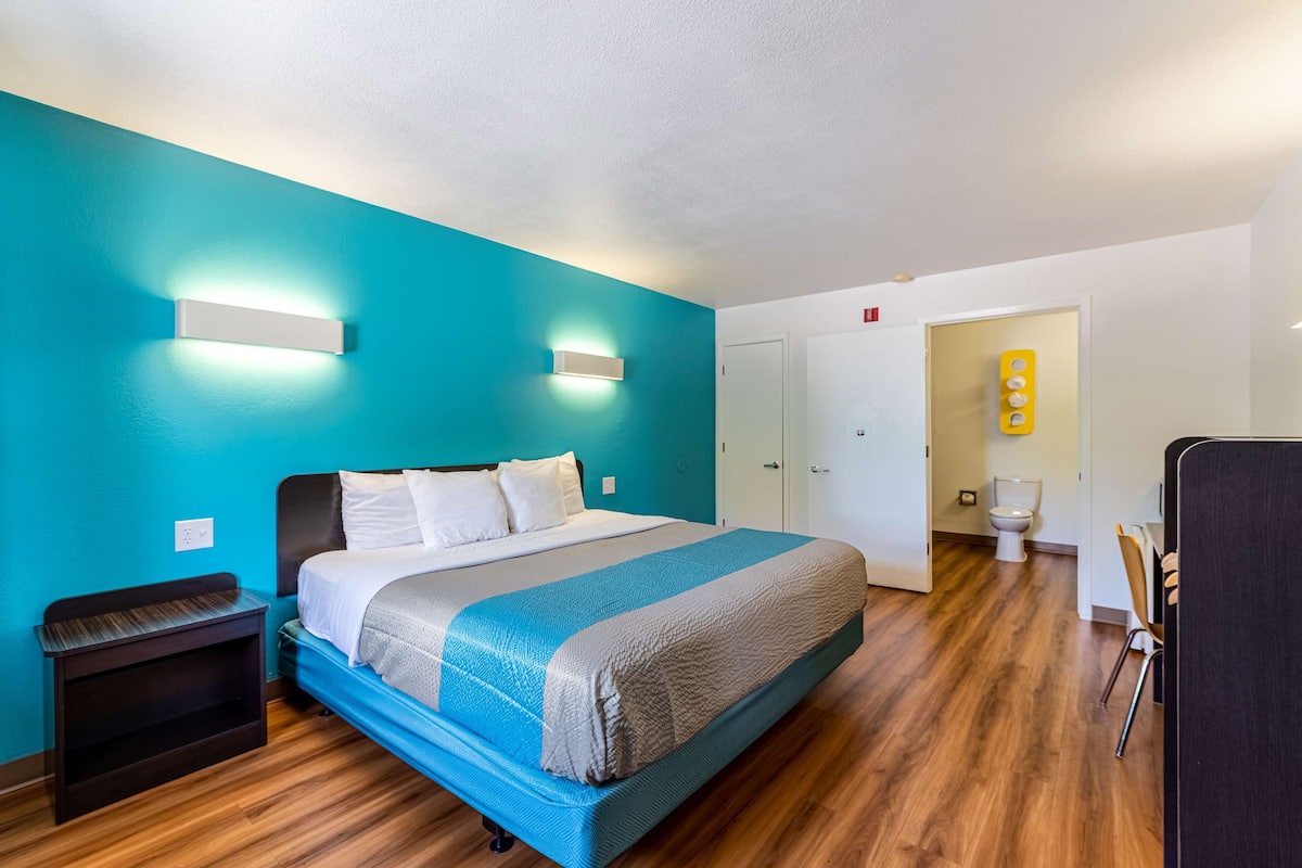 Quarto de hotel com uma cama, mesa de cabeceira, banheiro com vaso sanitário e porta que dá para outro quarto.