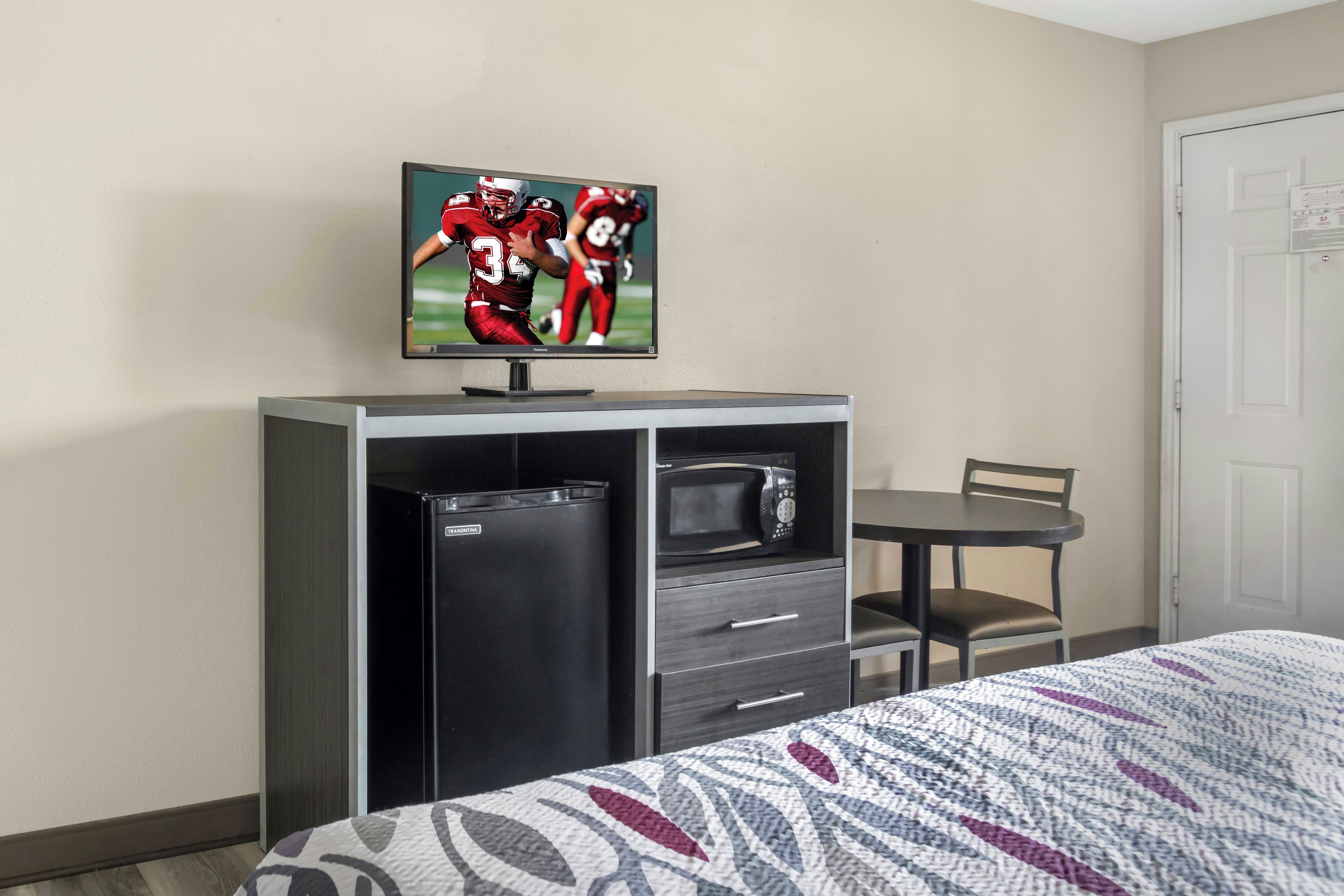 Quarto de hotel com cama, televisão presa em um móvel, frigorífico, micro-ondas e uma pequena mesa com cadeira.