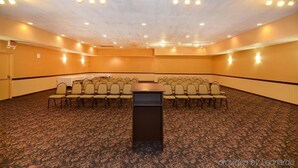 Uma sala de conferência com fileiras de cadeiras, uma mesa e piso coberto de carpete.