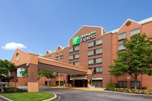 Um hotel Holiday Inn Express com fachada de tijolos, entrada coberta e área ajardinada com flores e árvores.