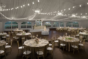Um amplo espaço para eventos coberto por uma tenda, com mesas redondas, cadeiras douradas e luzes de cordão.