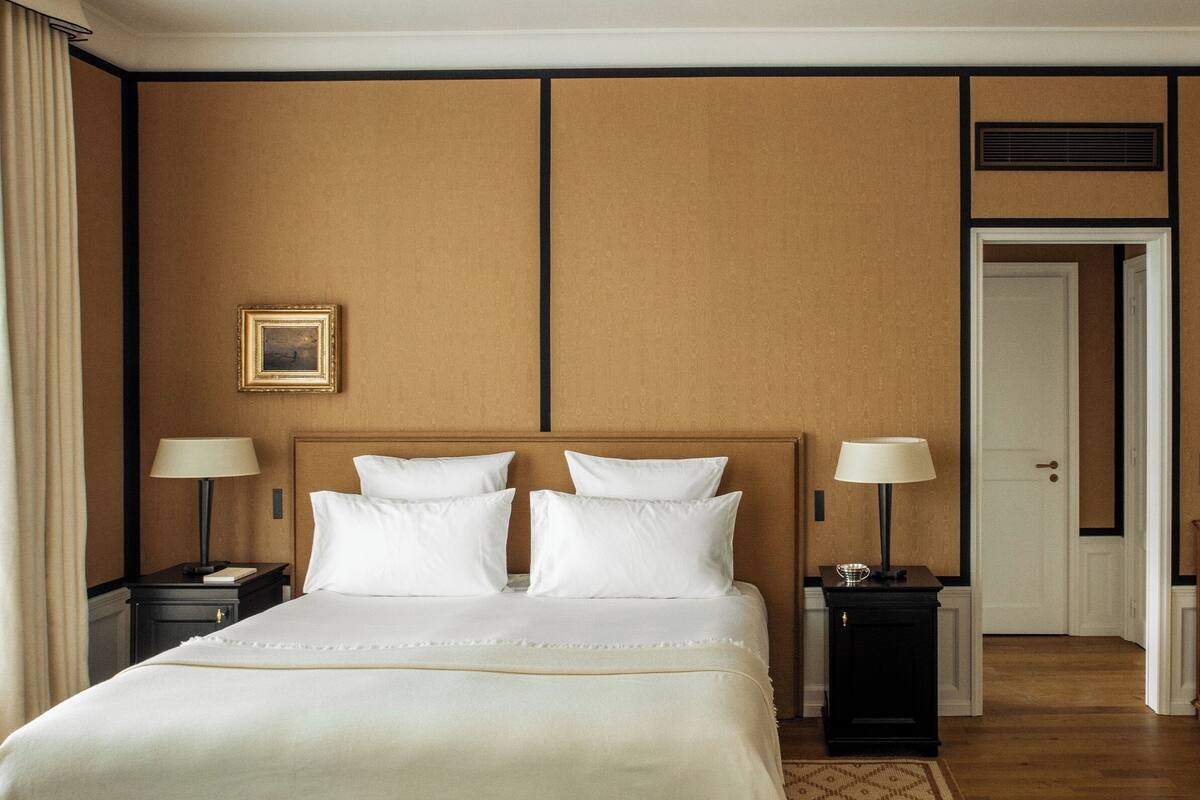 Quarto de hotel com uma cama grande, duas mesas de cabeceira com abajures, um quadro na parede e uma porta que dá para outro cômodo.