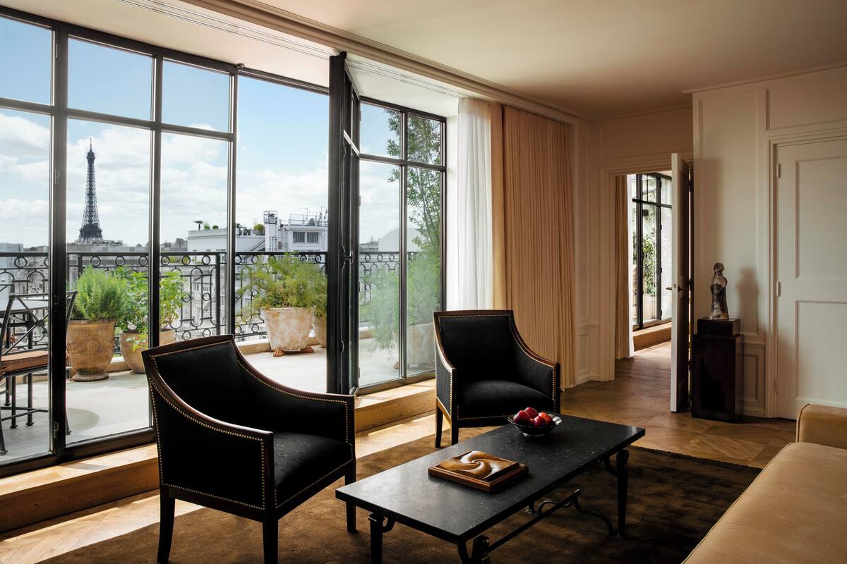 Quarto amplo com grandes janelas com vista para a Torre Eiffel, duas poltronas de couro preto, uma mesa de centro de madeira e uma pequena escultura em um pedestal.