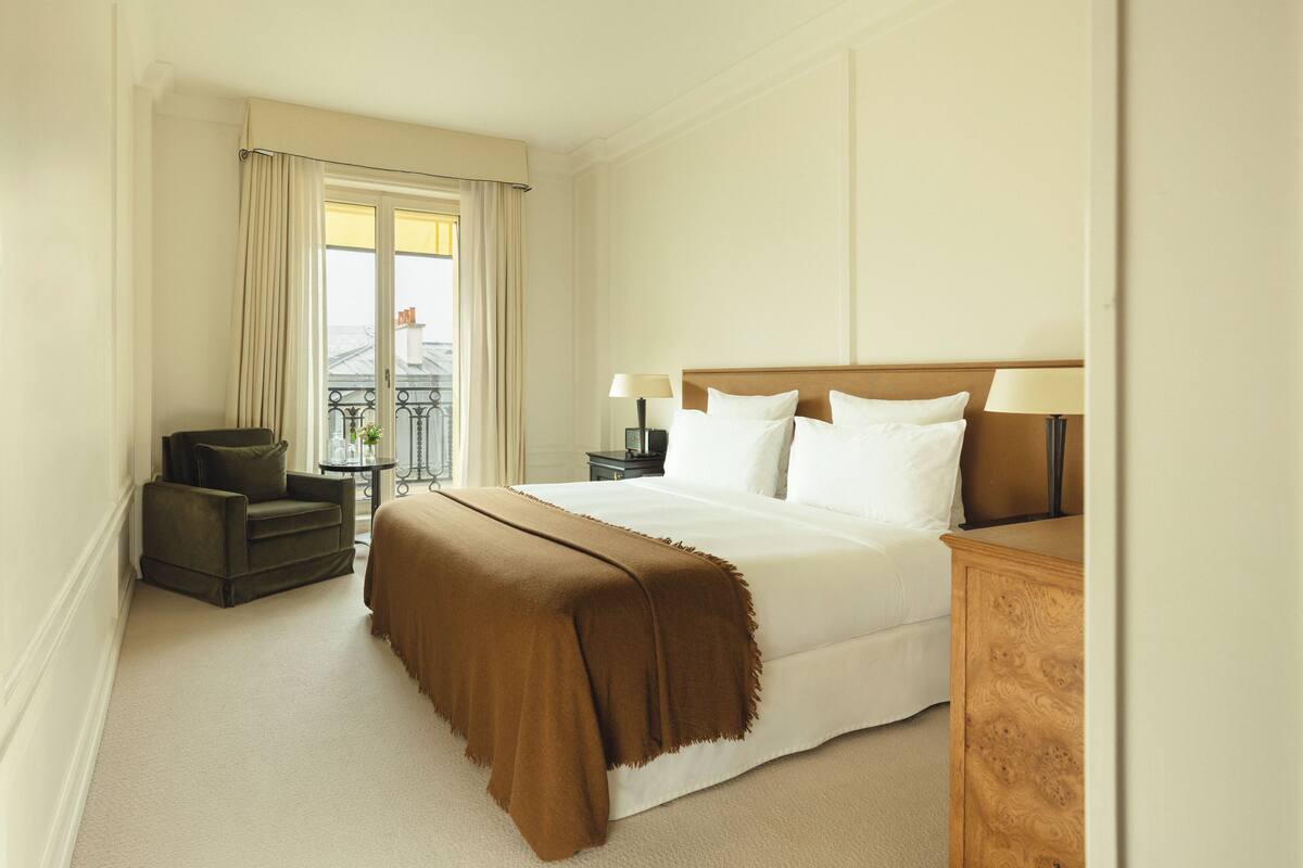 Quarto de hotel com uma cama grande, uma cadeira, uma mesa de cabeceira e vista para o exterior.