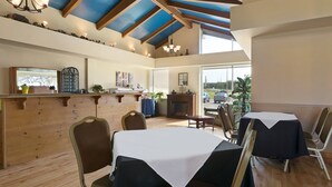 Uma área de refeições com piso de madeira, mesas com toalhas brancas e cadeiras. Há também um bar com balcão e objetos decorativos.
