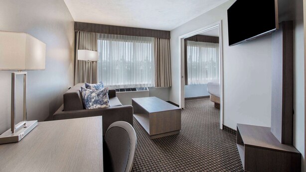 Quarto de hotel moderno com sofá, uma mesa pequena, televisão e uma janela grande com cortinas.