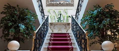 Uma escadaria imponente com tapete vermelho, ladeada por balaústres pretos e caneleiras brancas, que leva a um patamar com um quadro emoldurado.