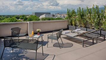 Terraço no telhado com área de estar ao ar livre, mesa com vaso e bebidas, e vista para a cidade e montanhas.