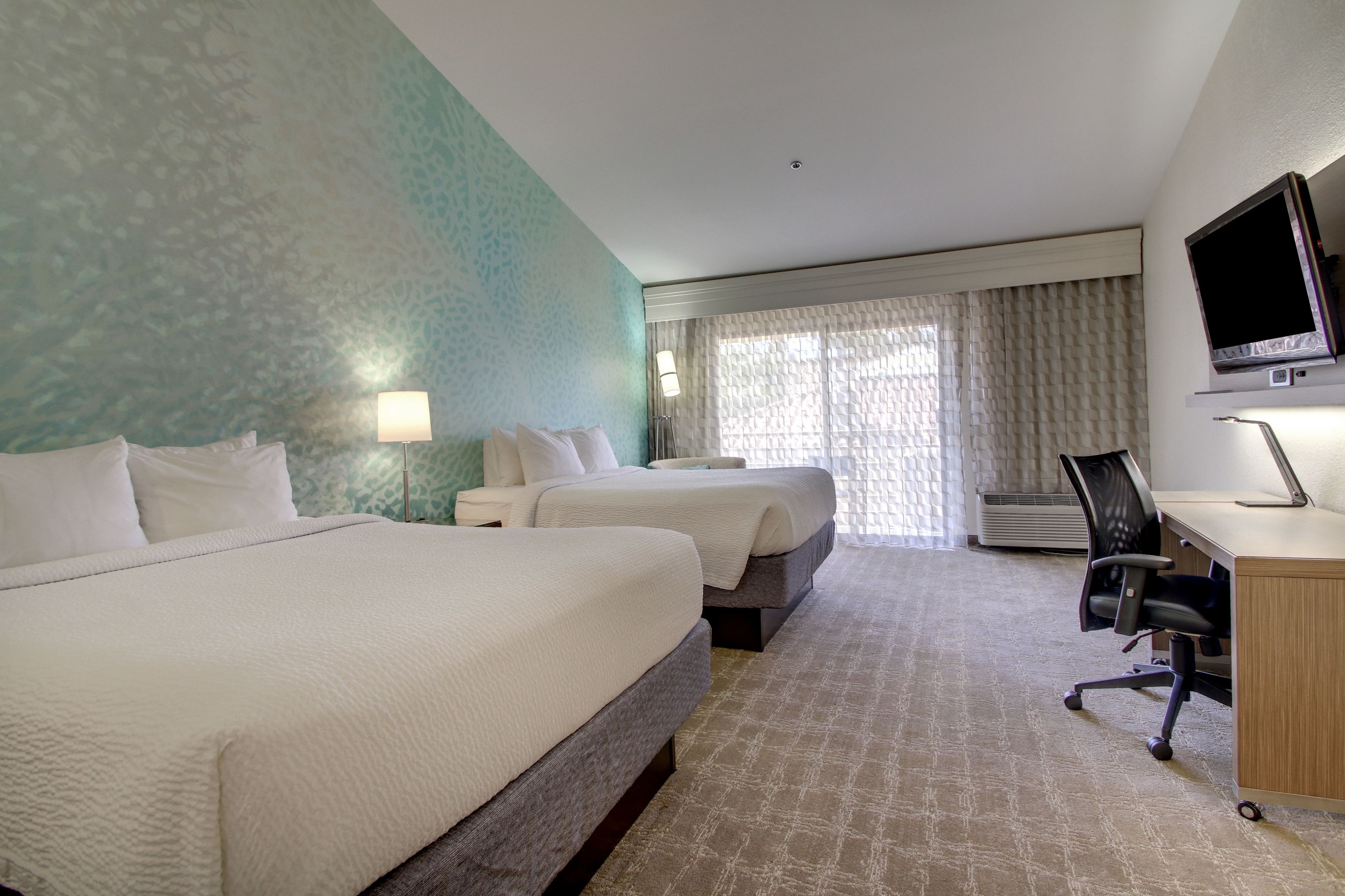 Quarto de hotel com duas camas, uma escrivaninha com cadeira, televisão e janela com cortinas.