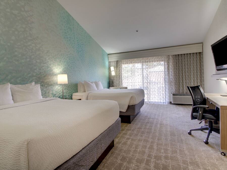 Quarto de hotel com duas camas, uma escrivaninha com cadeira, televisão e janela com cortinas.