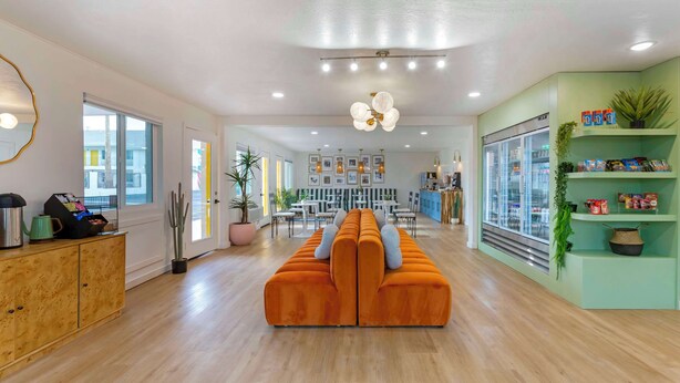 Uma sala de estar ampla com um sofá grande cor de laranja, piso de madeira e uma área de cozinha com geladeira e prateleiras.