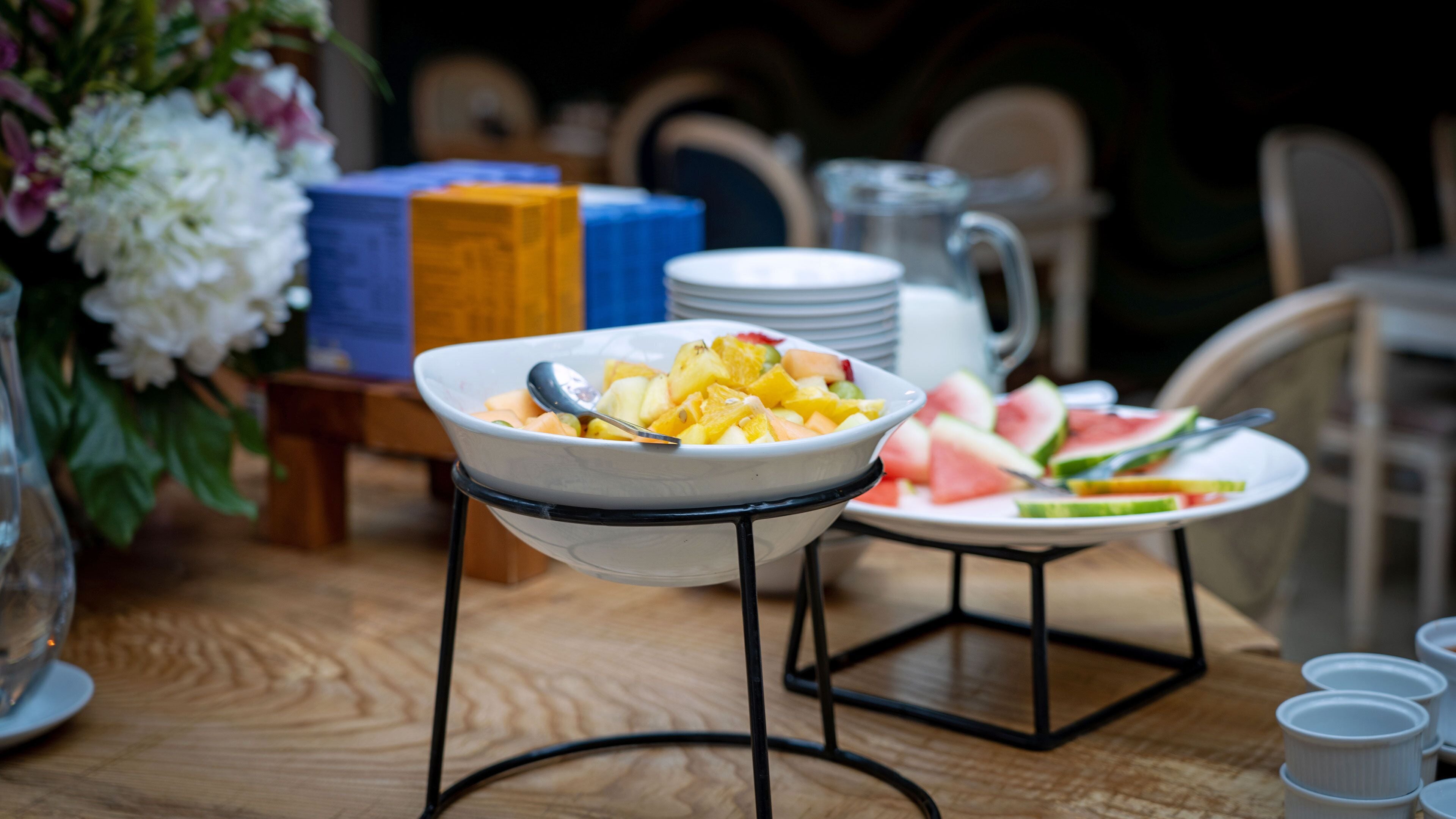 Uma mesa com uma fruteira, um prato de fatias de melancia e copos brancos empilhados.