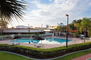 Piscina com espreguiçadeiras e guarda-sóis, cercada por uma vedação branca e vegetação.