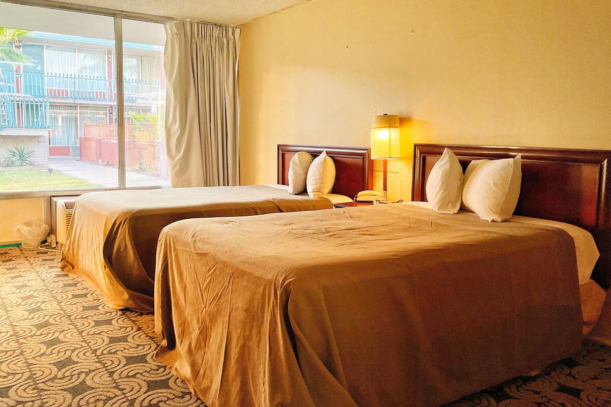 Quarto de hotel com duas camas, janela com cortinas, luminária e vista para o exterior.