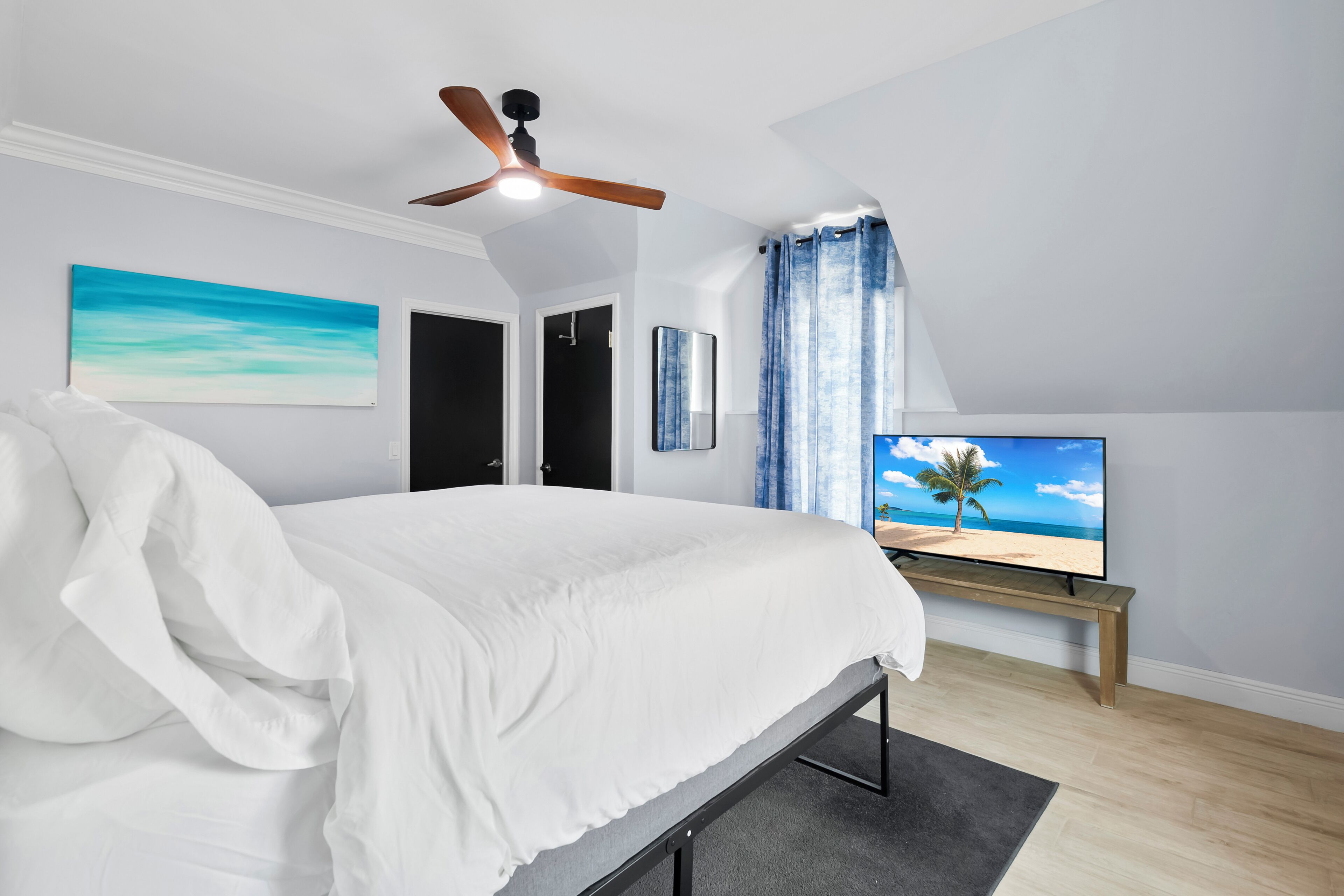 Quarto com uma cama, um ventilador de teto, uma TV exibindo uma cena de praia e um quadro de uma praia.
