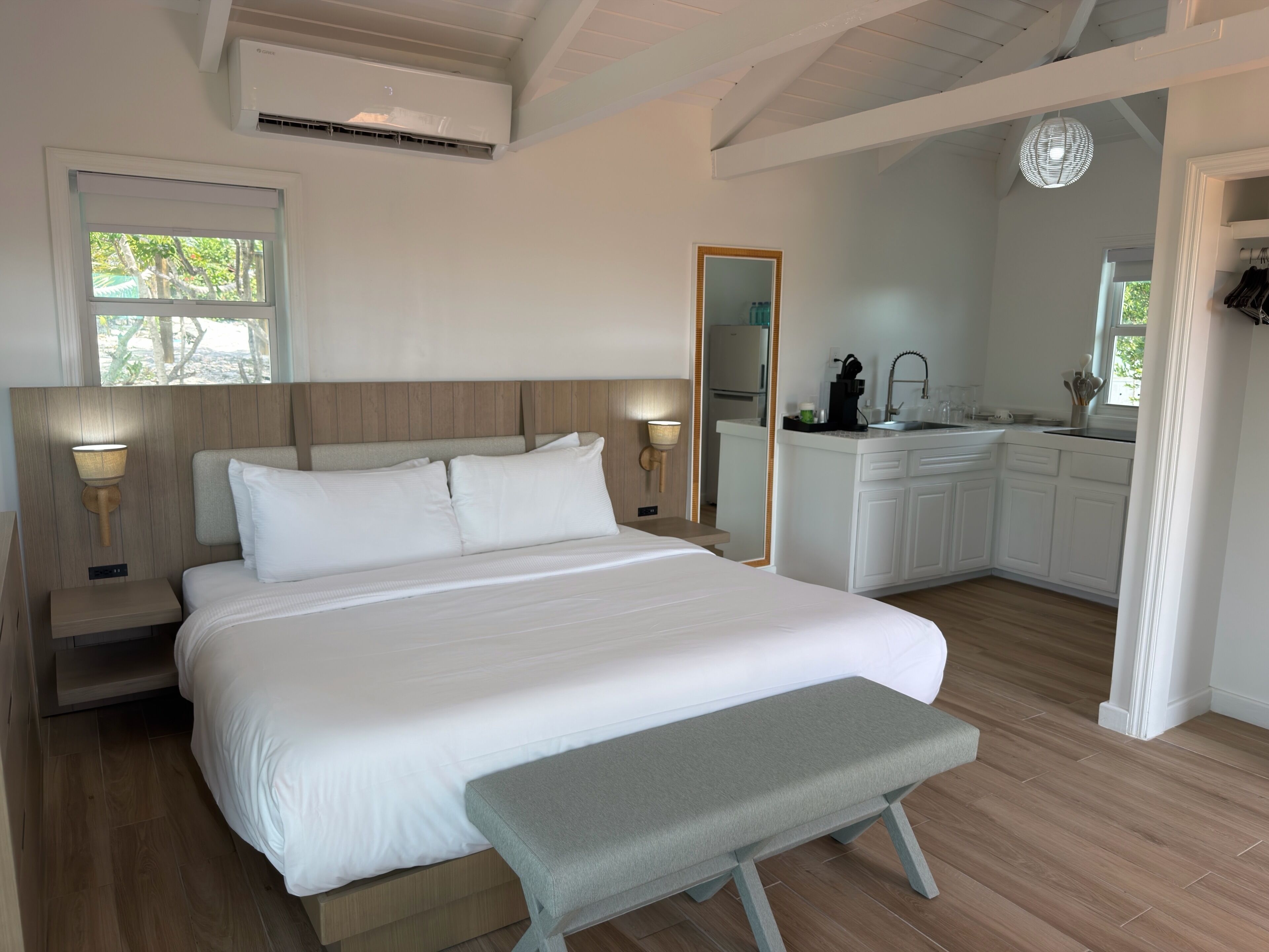Quarto de hotel com uma cama grande, luminárias de cabeceira, um banco, uma cozinha e um espelho.