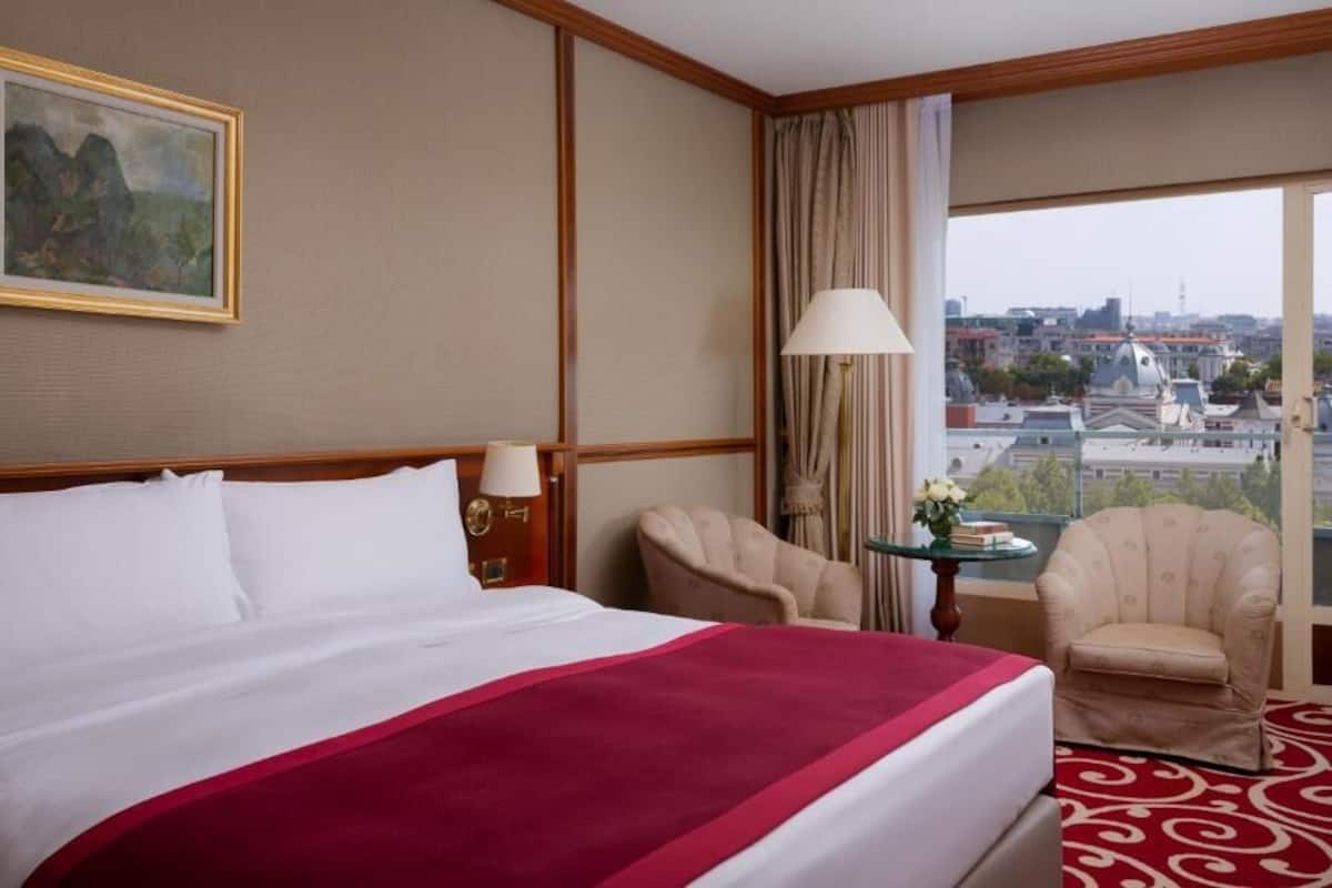 Quarto de hotel com uma cama grande, um quadro na parede, vista da cidade pela janela e uma cadeira junto à janela.
