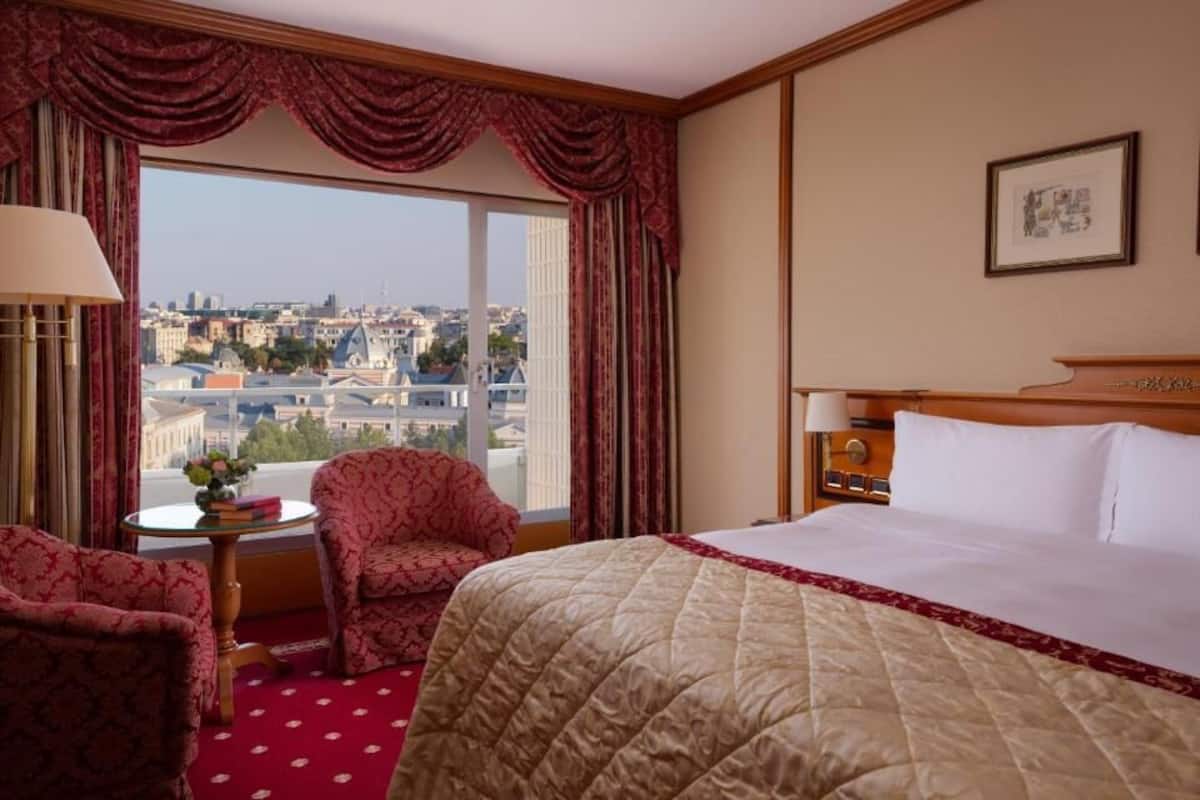 Quarto de hotel com uma cama grande, uma cadeira, uma mesa pequena com um vaso e vista da cidade através de uma janela ampla.