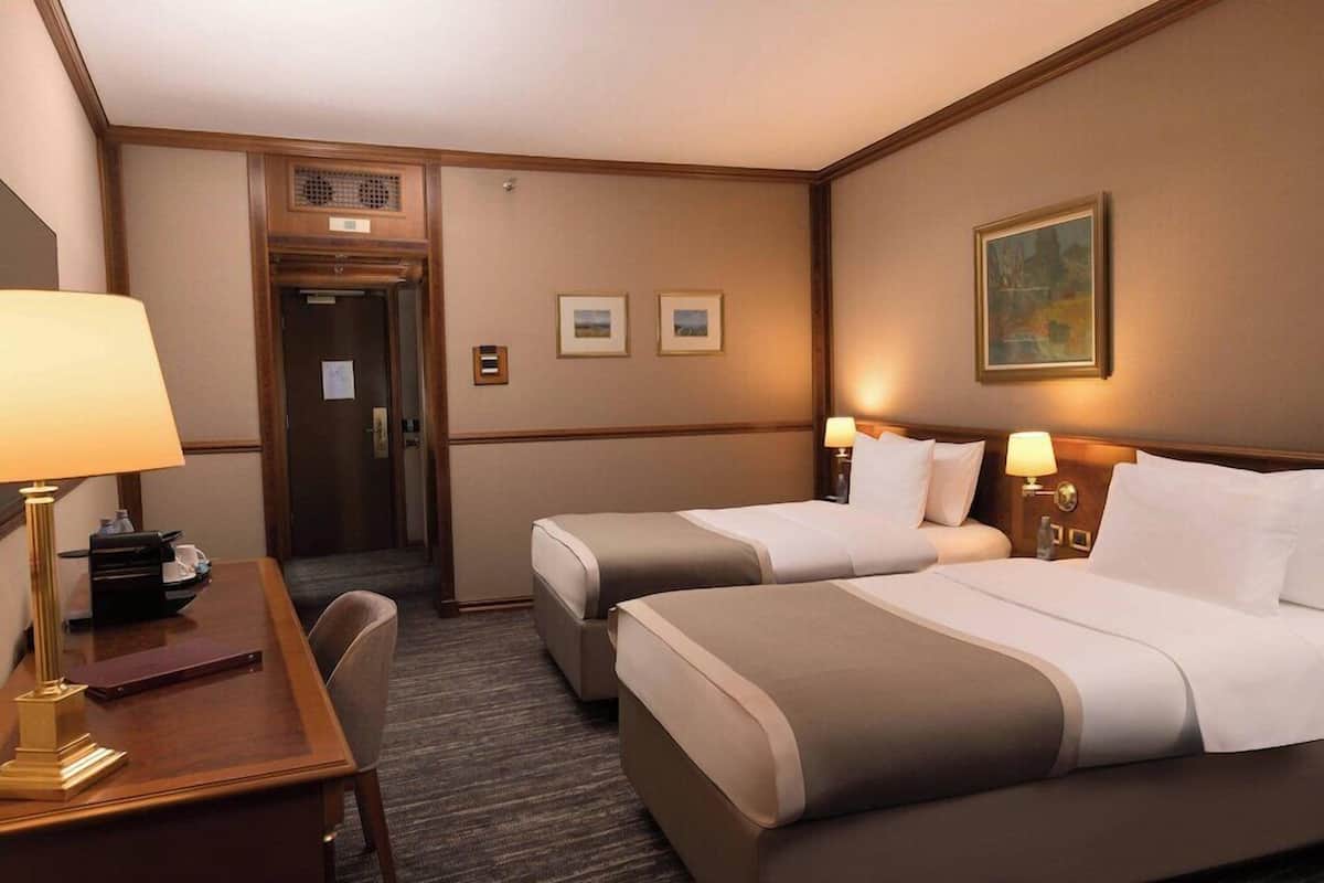 Quarto de hotel com duas camas, uma escrivaninha com abajur, uma cadeira e uma porta que dá para um corredor.