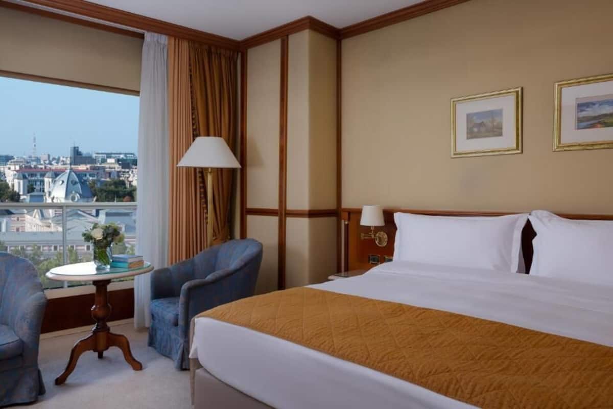 Quarto de hotel com uma cama grande, duas poltronas, uma mesinha com um vaso, uma luminária e quadros nas paredes.