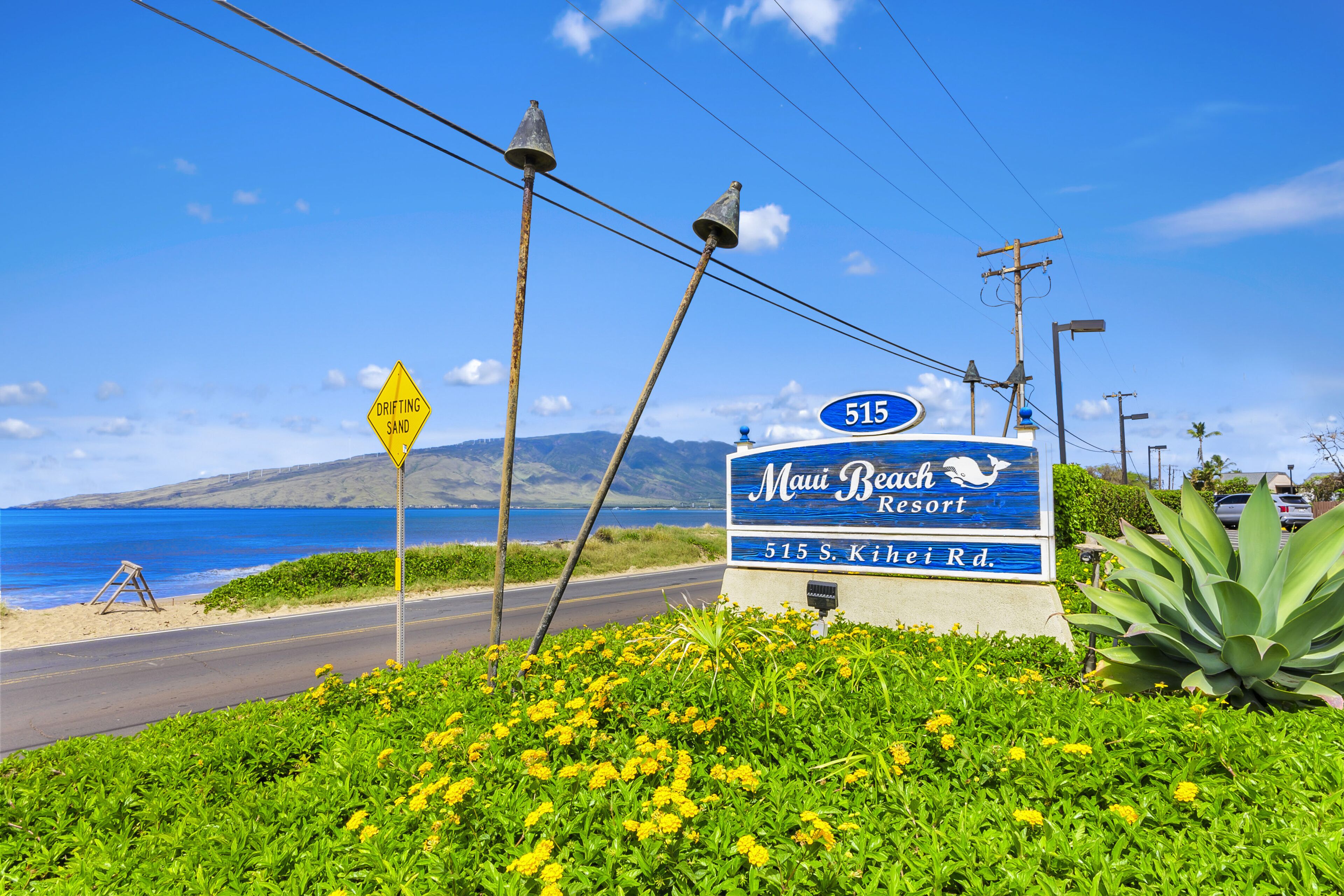 Placa do Maui Beach Resort com vista para a praia, uma placa amarela com a inscrição 'Drifting Sand' e um céu azul claro.