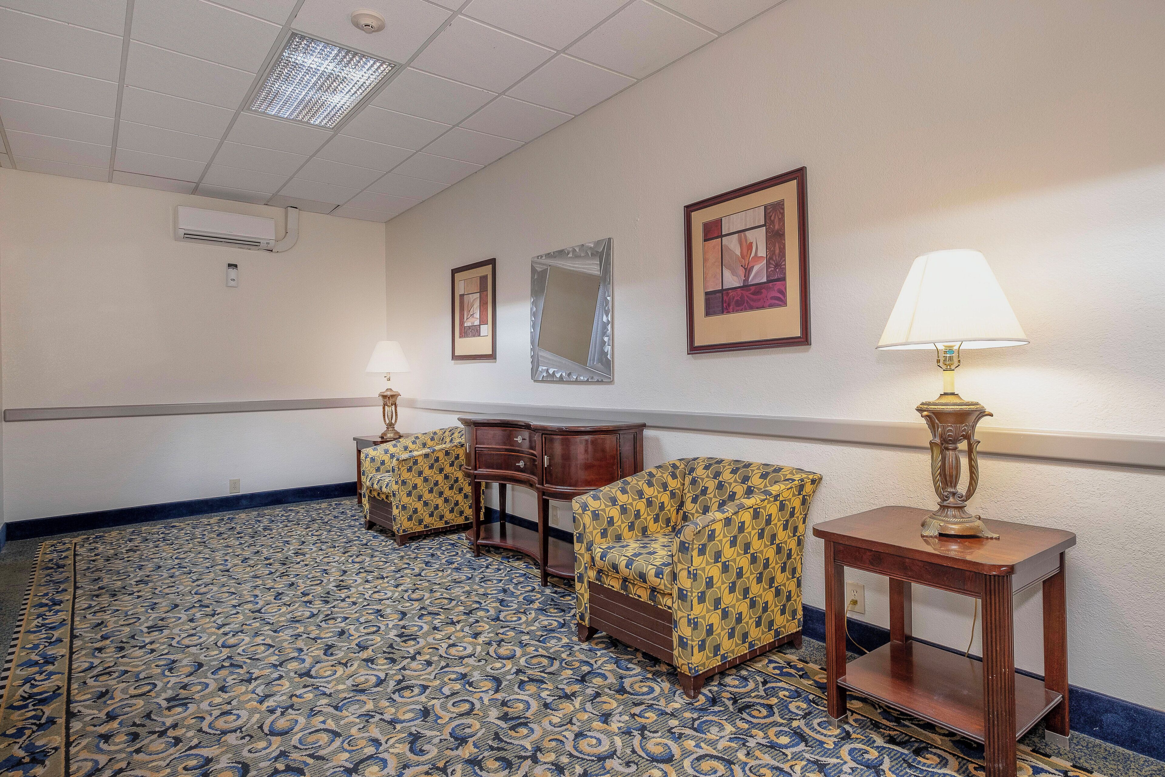 Quarto de hotel com carpete estampado, mesa de cabeceira de madeira com abajur, cadeira de madeira, pequeno armário de madeira e espelho fixado na parede.
