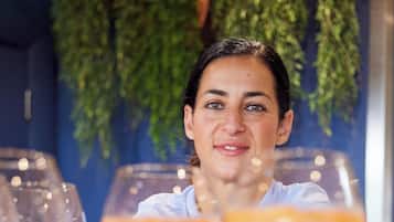Uma pessoa segurando dois copos de vinho em uma mesa, com uma parede azul e vegetação pendente ao fundo.