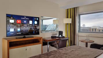 Quarto de hotel com televisão exibindo diversos canais, uma escrivaninha com cadeira, uma mesinha de cabeceira com abajur e uma janela com vista para a cidade.